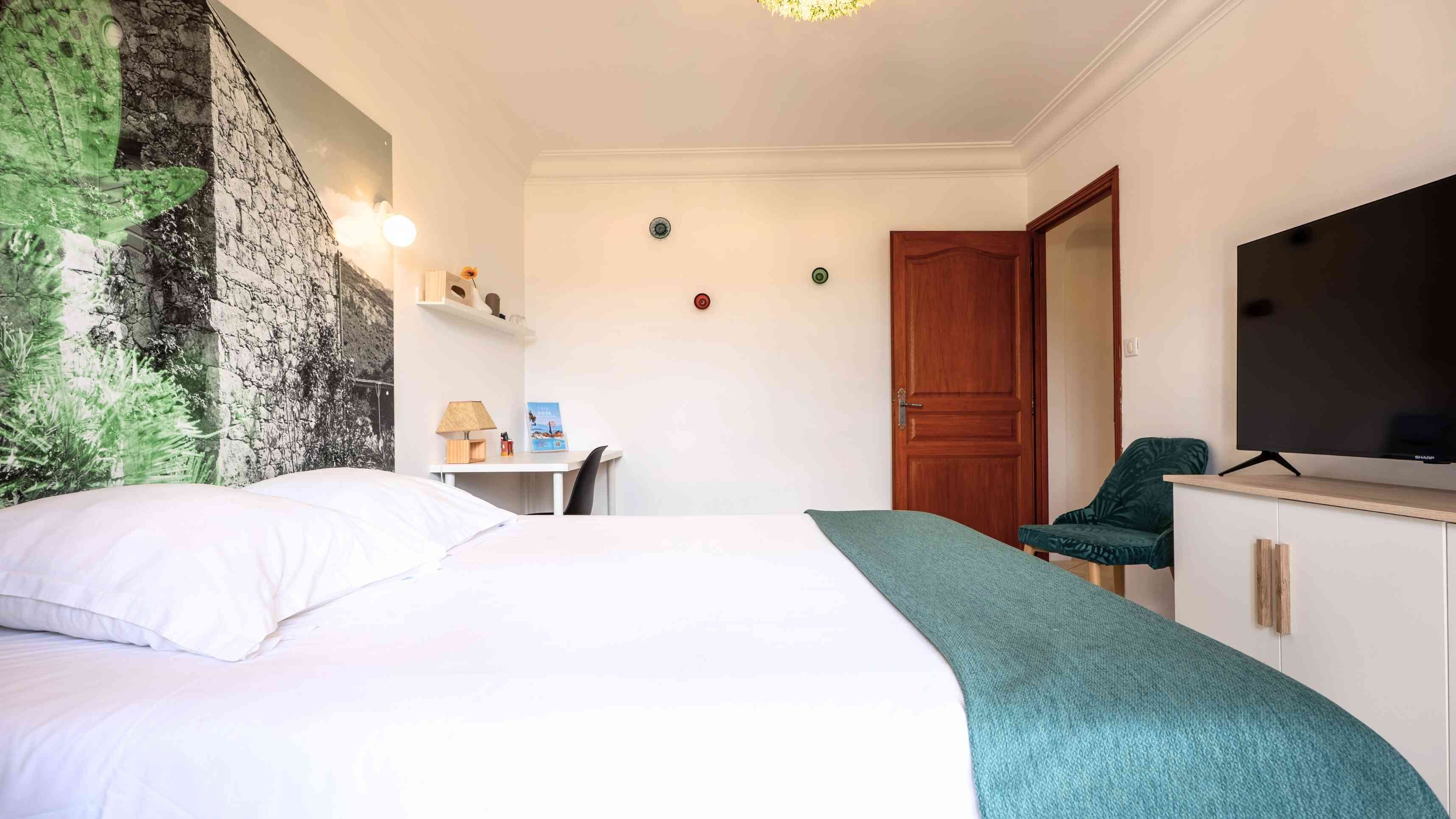 Quarto de hotel com cama, escrivaninha, cadeira, televisão e guarda-roupa.