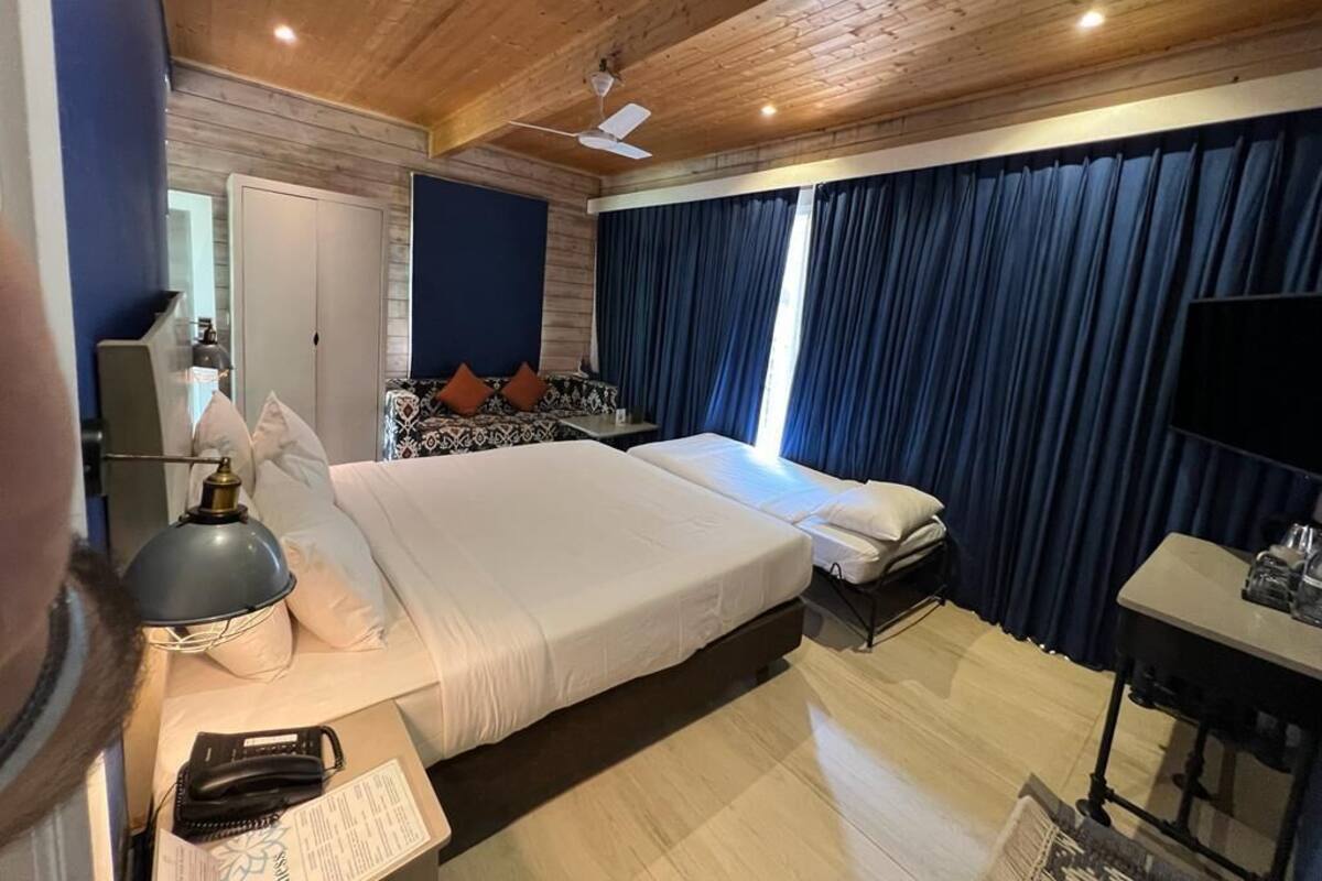 Uma cama de casal com lençóis brancos, uma escrivaninha com abajur, um telefone e uma mesinha com uma garrafa.