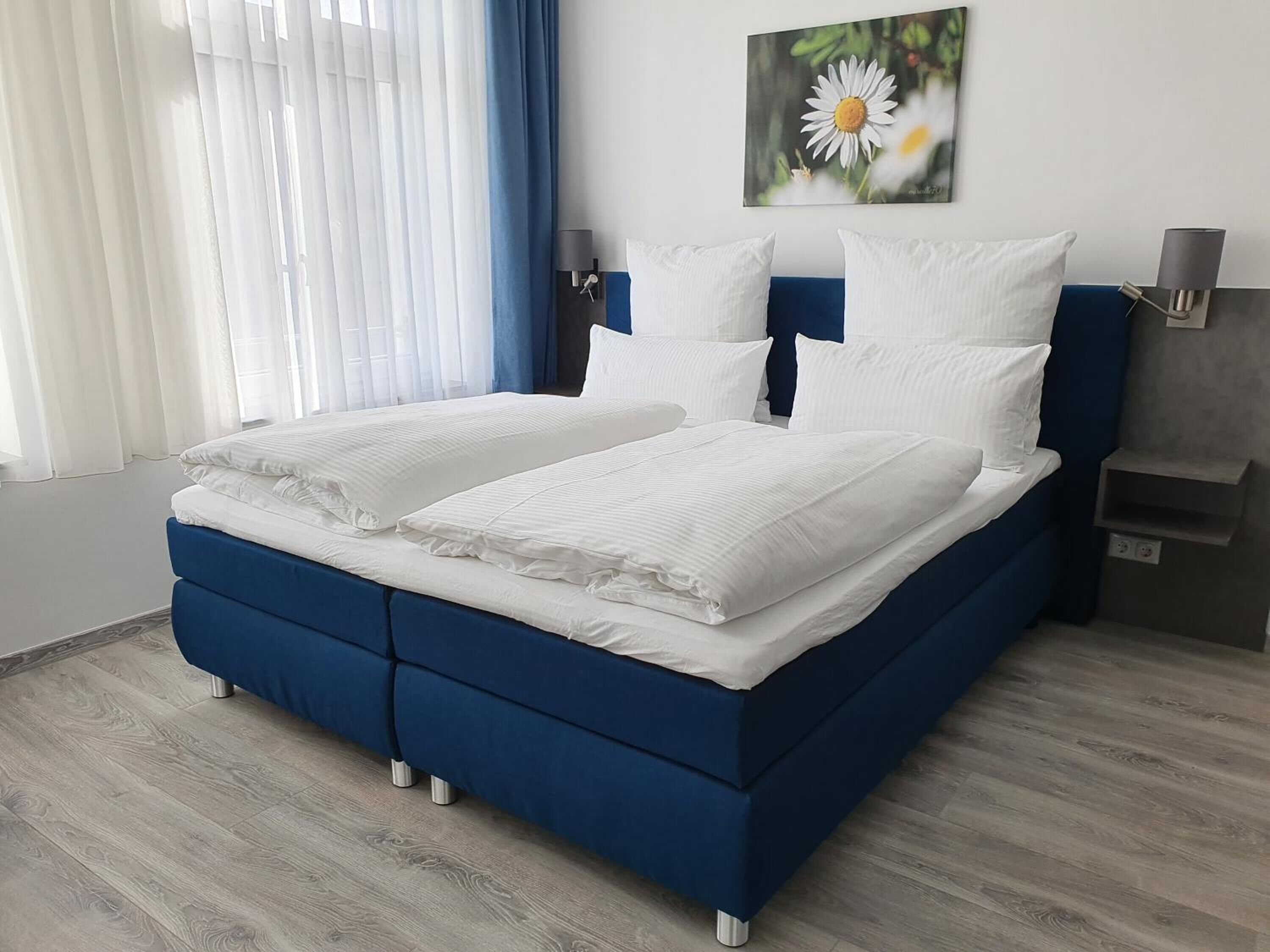 Quarto moderno com cama azul, roupa de cama branca e uma luminária fixada na parede.