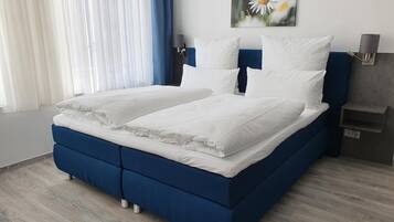 Quarto moderno com cama azul, roupa de cama branca e uma luminária fixada na parede.
