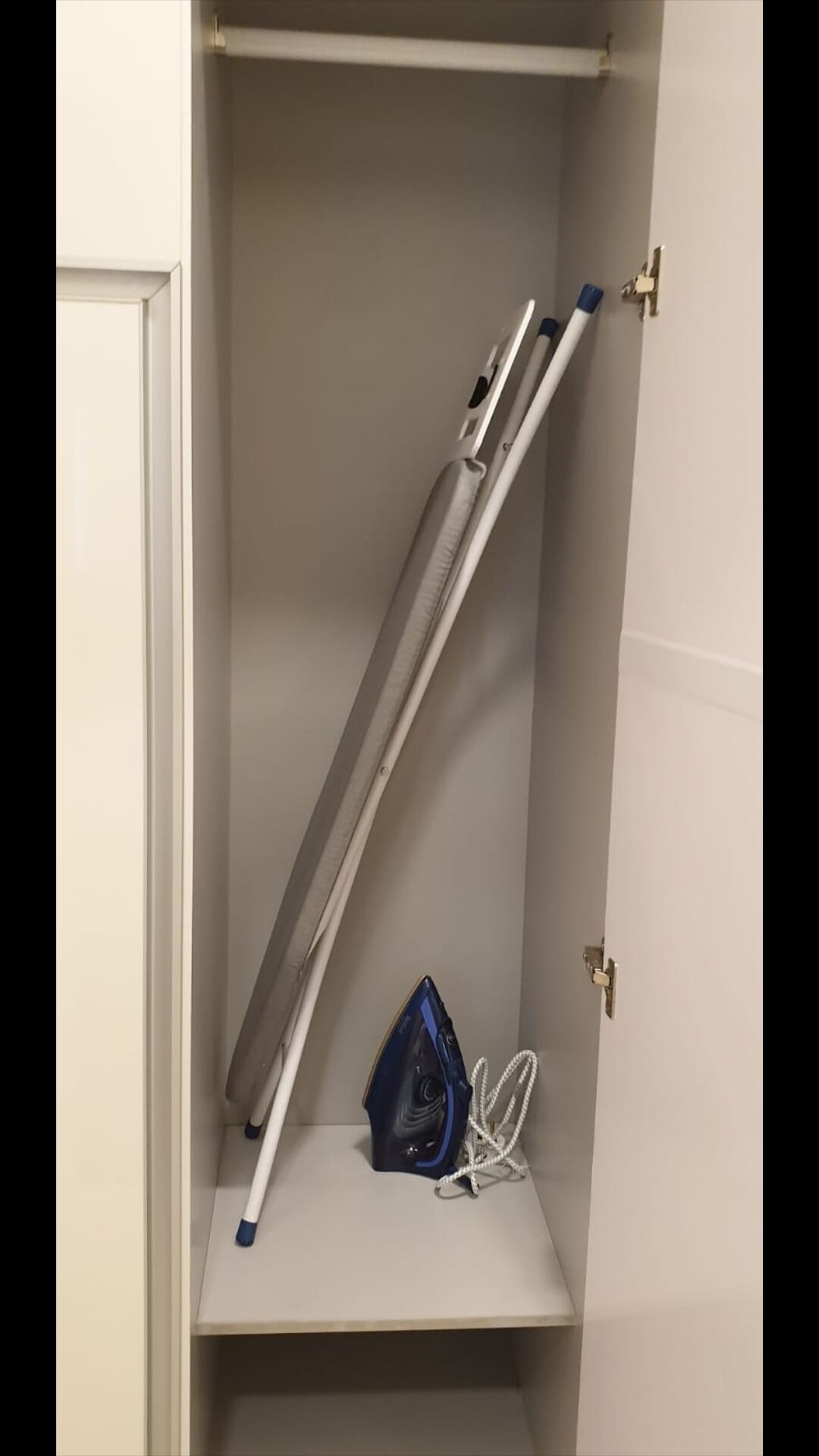 Armário com barra para pendurar roupas e prateleira contendo um ferro azul e um cabo branco.