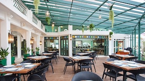 Restaurante acolhedor com mesas e cadeiras, teto de vidro e plantas pendentes.