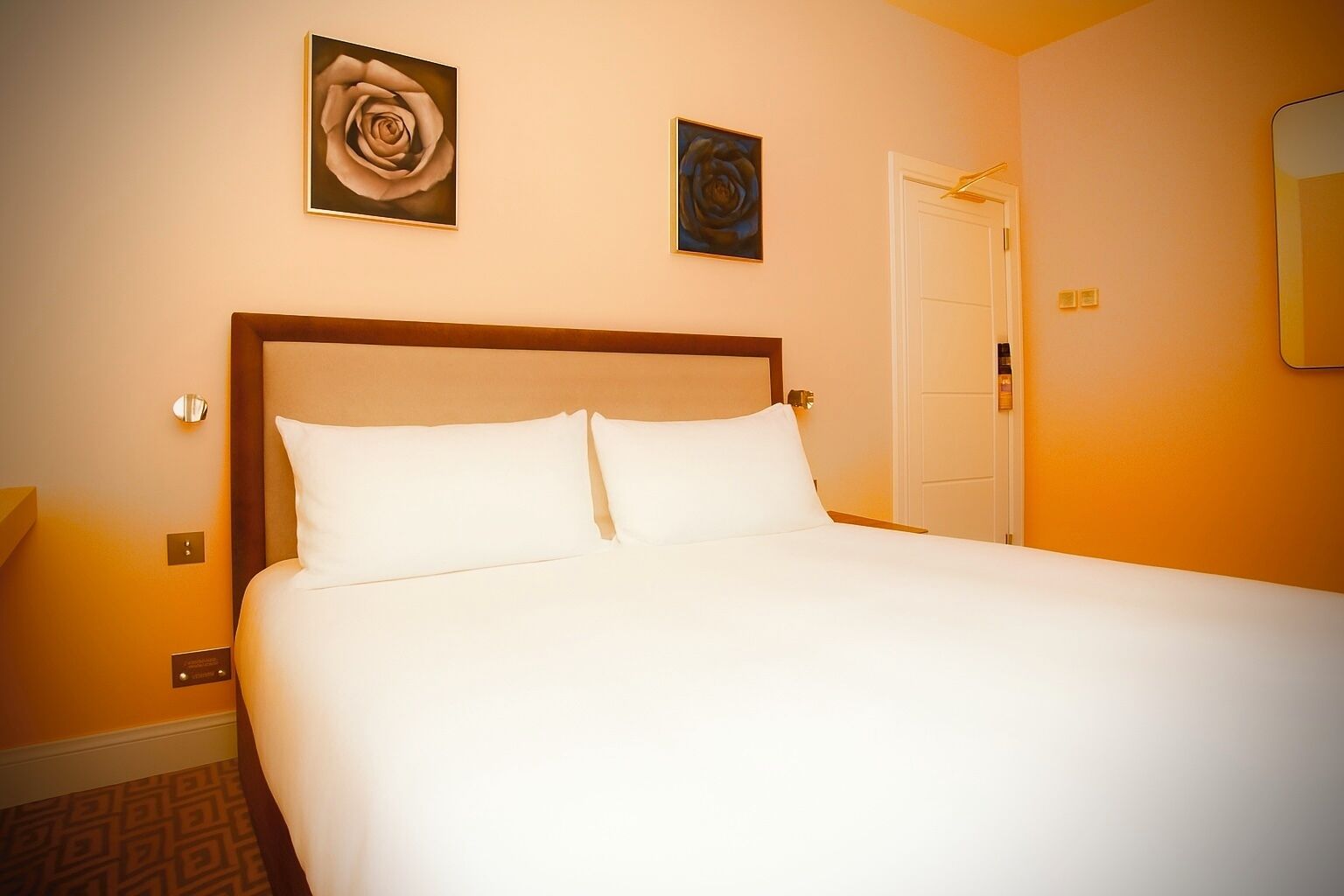 Quarto de hotel com uma cama grande, duas pinturas penduradas na parede, uma porta e um interruptor de luz.