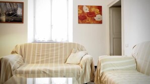 Uma sala de estar com um sofá e uma poltrona, uma janela com cortinas e duas pinturas nas paredes.