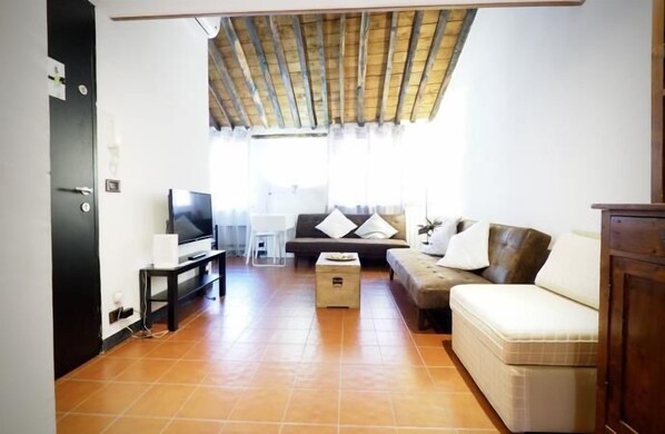 Sala de estar com televisão, sofá e apoio para os pés. Teto de madeira e janela com cortinas.