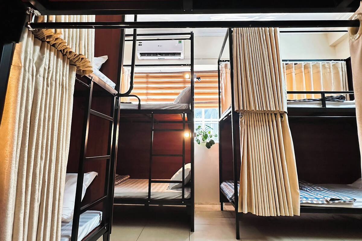 Quarto em dormitório com beliches, janela com persianas, ar condicionado e pequena área de estudo.