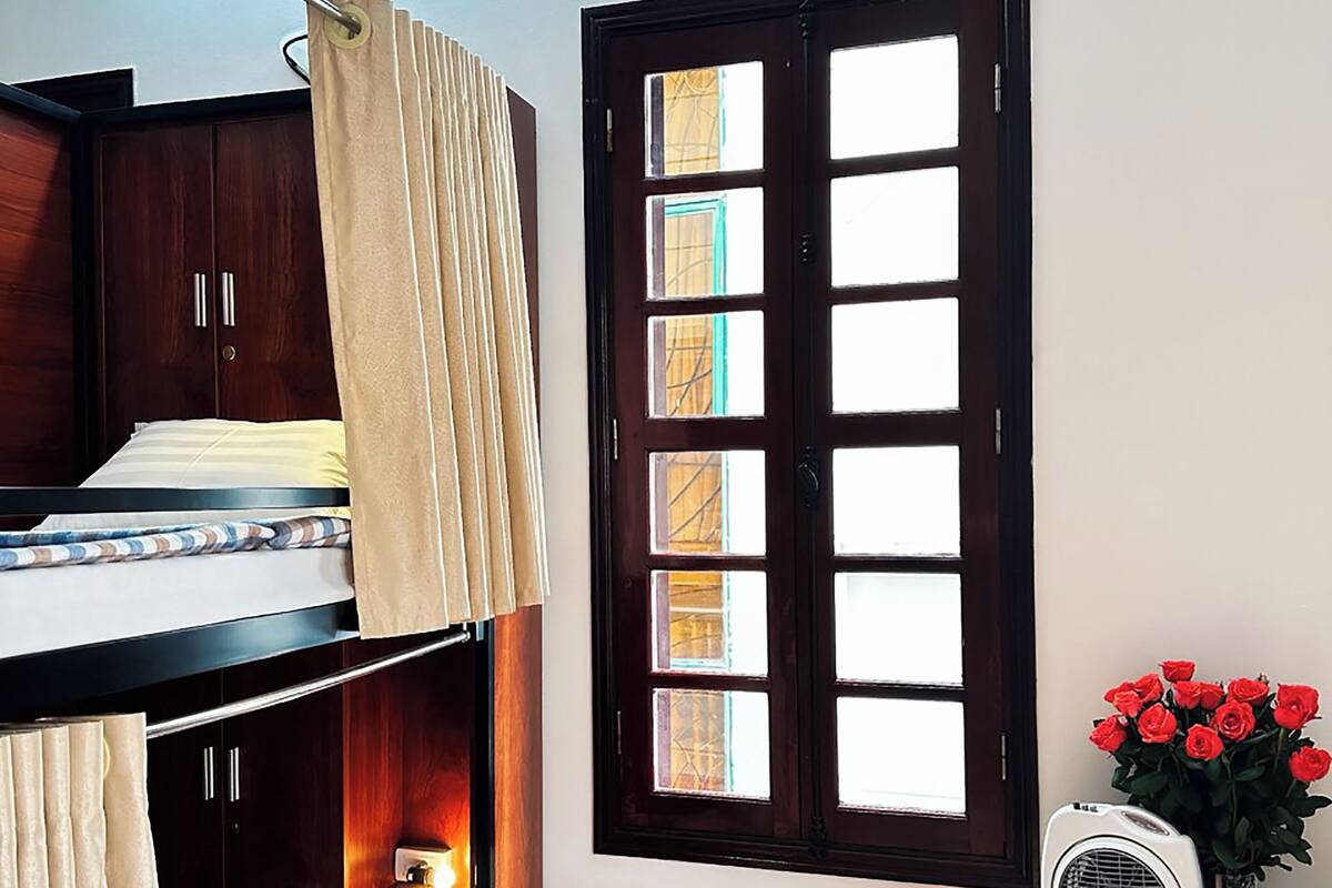 Um dormitório com uma cama em beliche, uma janela com vários painéis, uma mesa de cabeceira com um ventilador e um vaso de flores vermelhas.