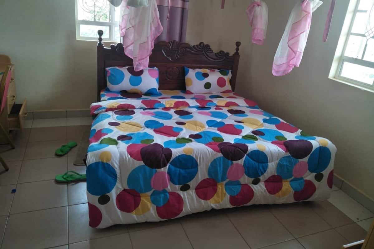 Uma cama com colcha estampada em bolinhas coloridas, cabeceira de madeira e janela com cortinas.