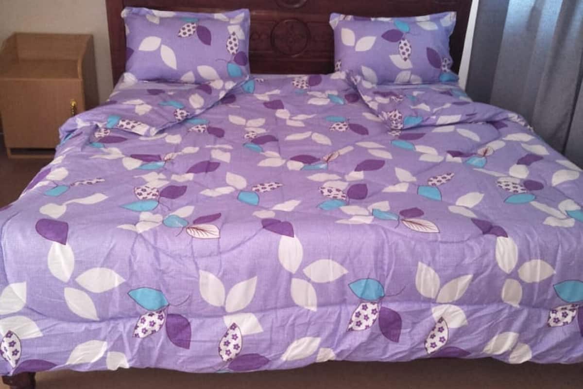 Uma cama com colcha e almofadas florais lilases, uma cabeceira de madeira e uma mesa de cabeceira à esquerda.