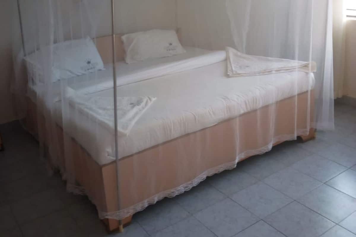 Uma cama com dossel e cortina, com um edredom branco e almofadas, em um quarto com piso de azulejos e parede lisa.