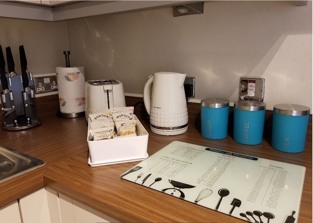 Uma bancada de cozinha com cafeteira, recipientes de chá e café, uma caixa de cápsulas de café, torradeira e um cardápio.