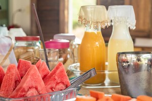 Uma mesa com diversos alimentos e bebidas, incluindo melancia, um jarro de suco de laranja e uma garrafa de bebida branca.