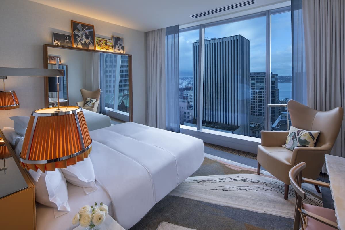 Quarto de hotel moderno com uma cama grande, um espaço de estar com uma poltrona, uma mesa e vista para a cidade através de amplas janelas.