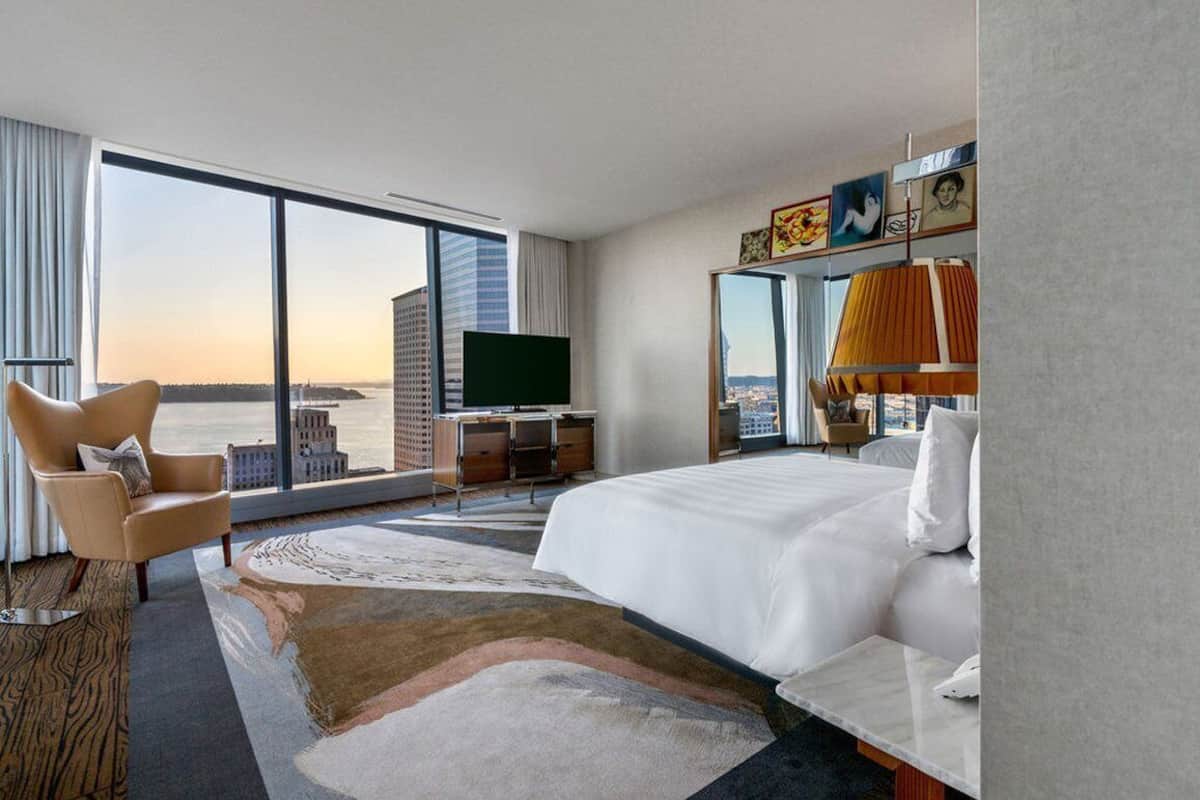 Quarto de hotel com uma cama grande, televisão, escrivaninha e cadeira. Há uma janela ampla com vista para a cidade.