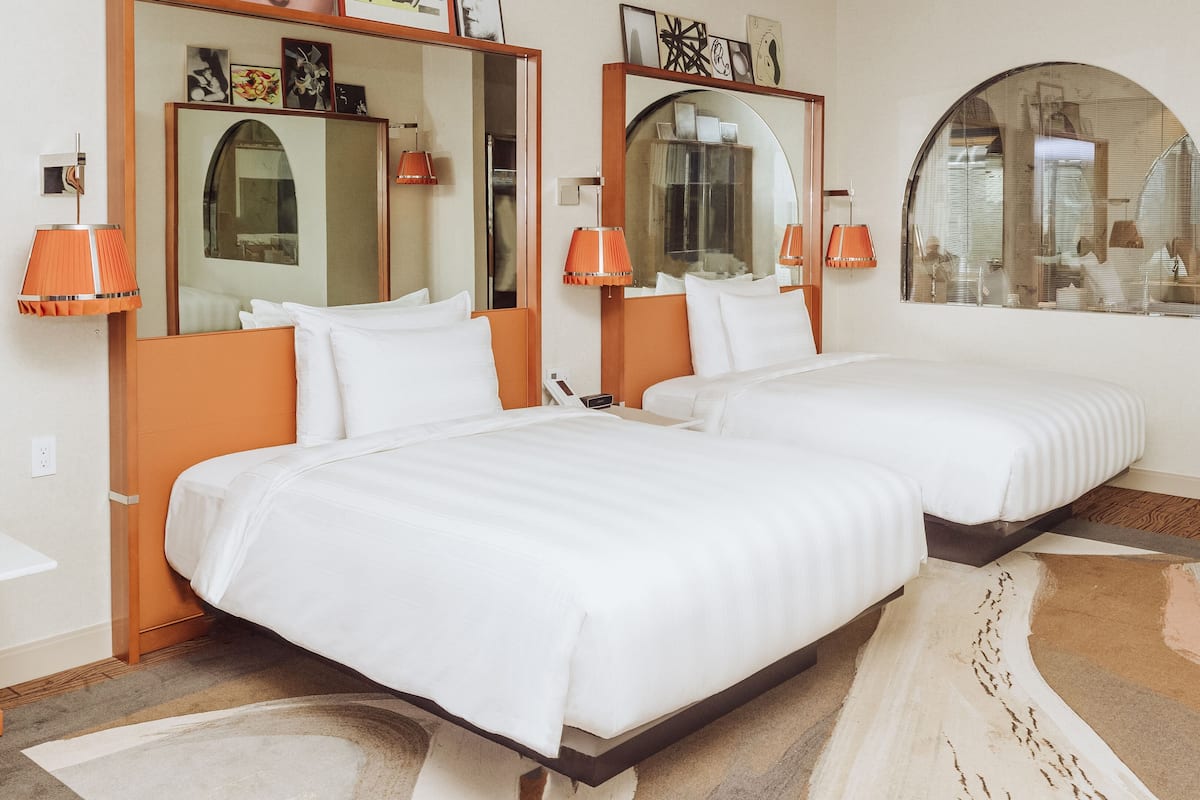 Quarto de hotel com duas camas, um espelho grande, uma mesa pequena e um tapete decorativo.