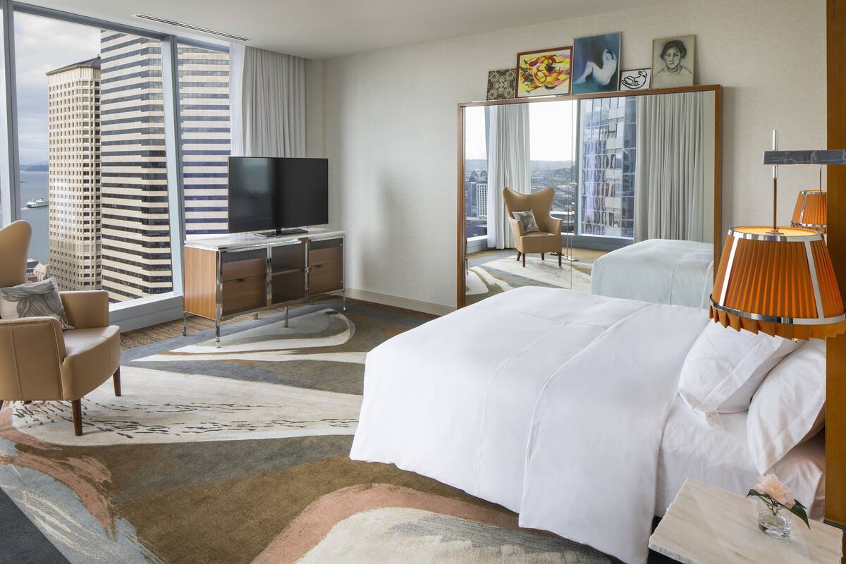 Quarto de hotel com uma cama grande, televisão, secretária e uma cadeira. Vê-se edifícios pela janela.