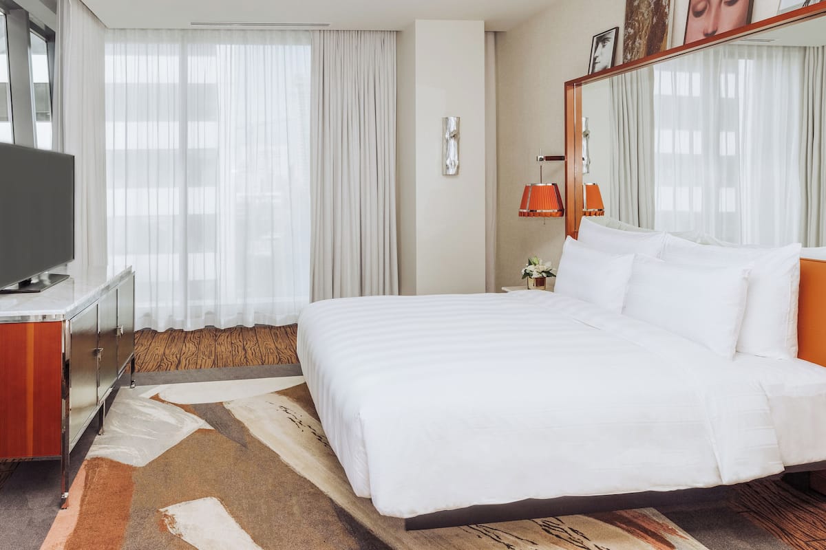 Quarto de hotel com uma cama grande, uma televisão sobre um suporte, uma mesa de cabeceira de madeira e um espelho.