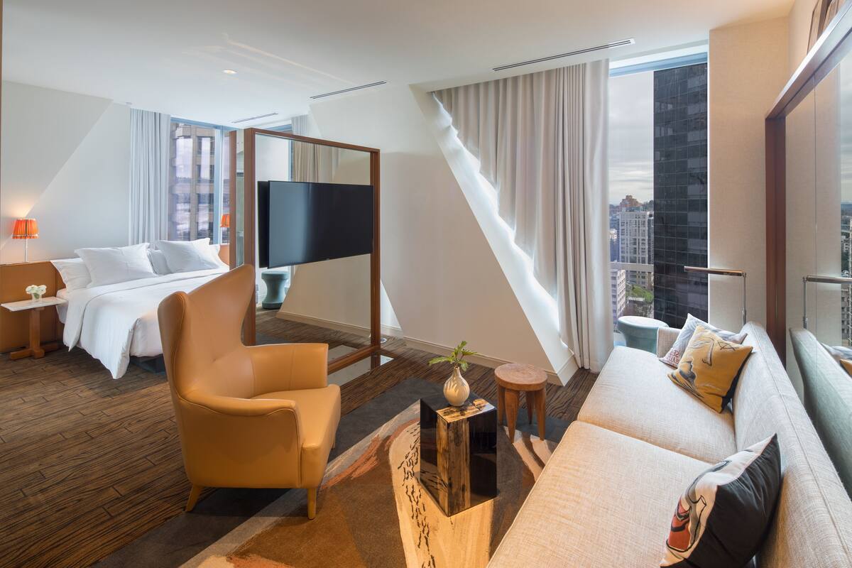 Quarto de hotel moderno com cama, sofá, poltrona, TV e varanda com cortinas.