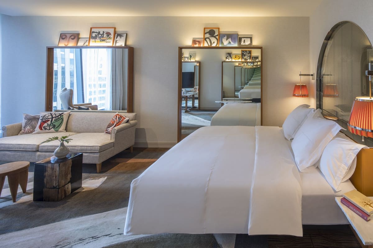 Quarto de hotel com uma cama grande, um sofá, uma pequena mesa de madeira e um espelho.