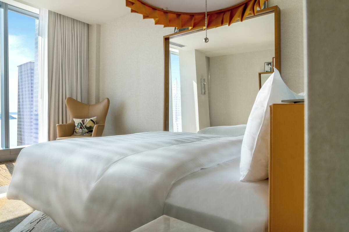 Quarto de hotel com uma cama grande, uma cadeira, uma escrivaninha e uma janela ampla com vista para a cidade.