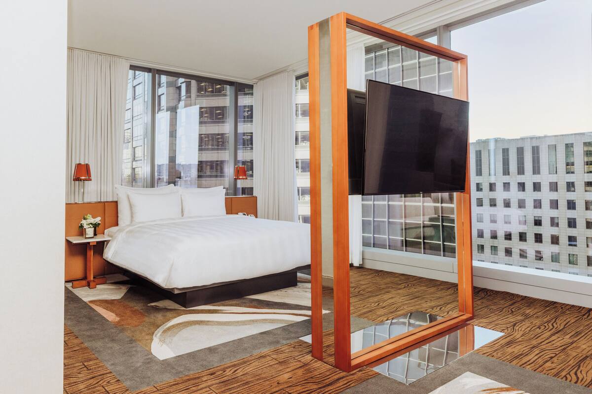 Quarto de hotel moderno com uma cama grande, televisão e vista para a paisagem urbana.