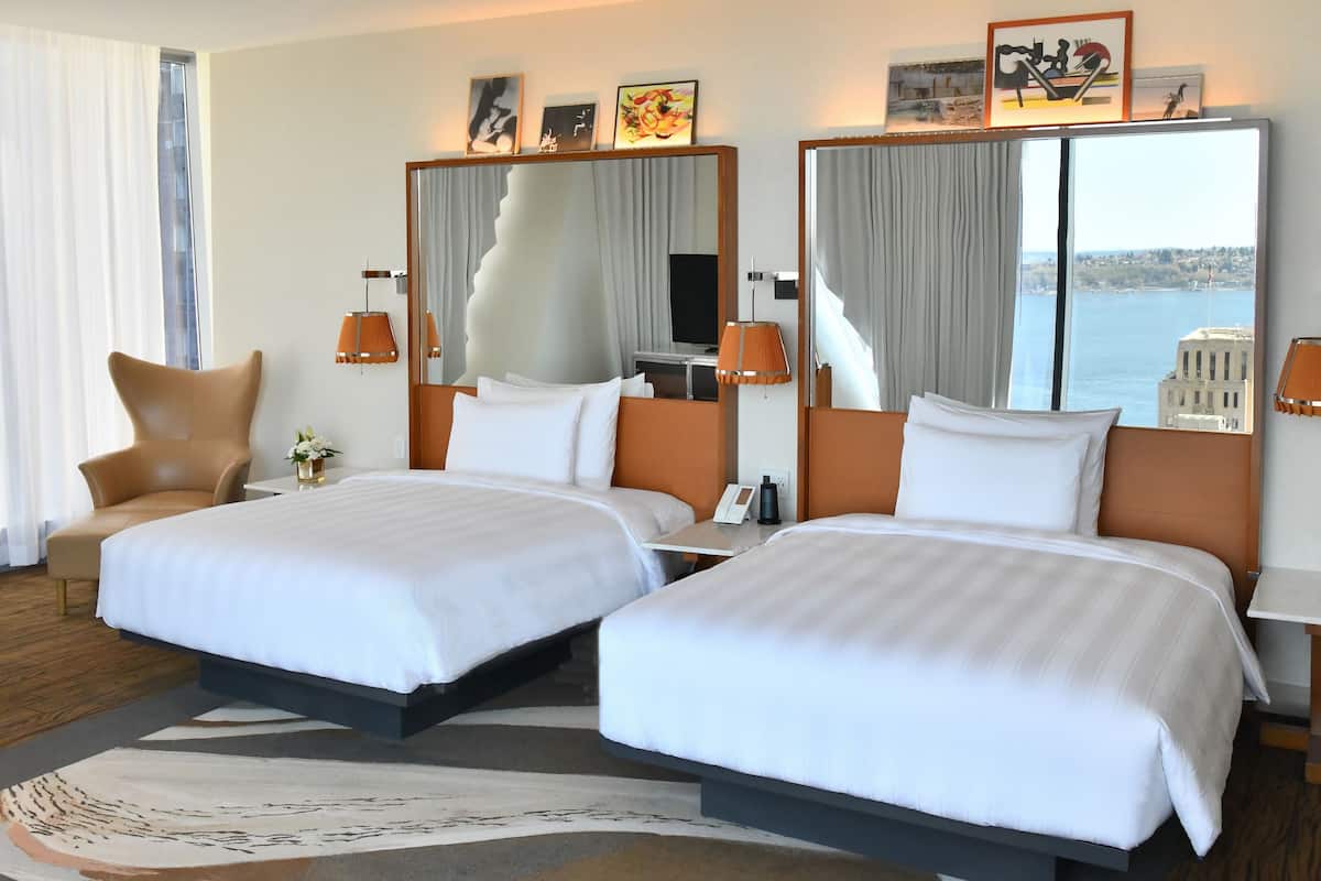 Quarto de hotel com duas camas, uma cadeira, uma escrivaninha e vista para o mar.