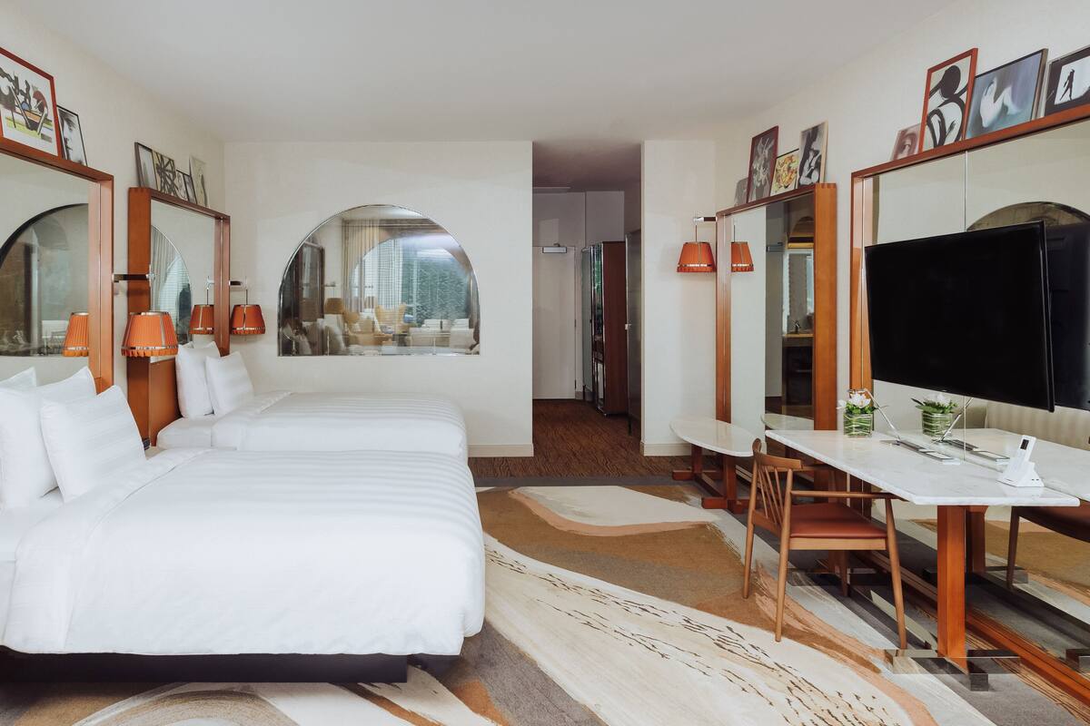 Quarto de hotel com duas camas, uma escrivaninha com televisão e obras de arte nas paredes.