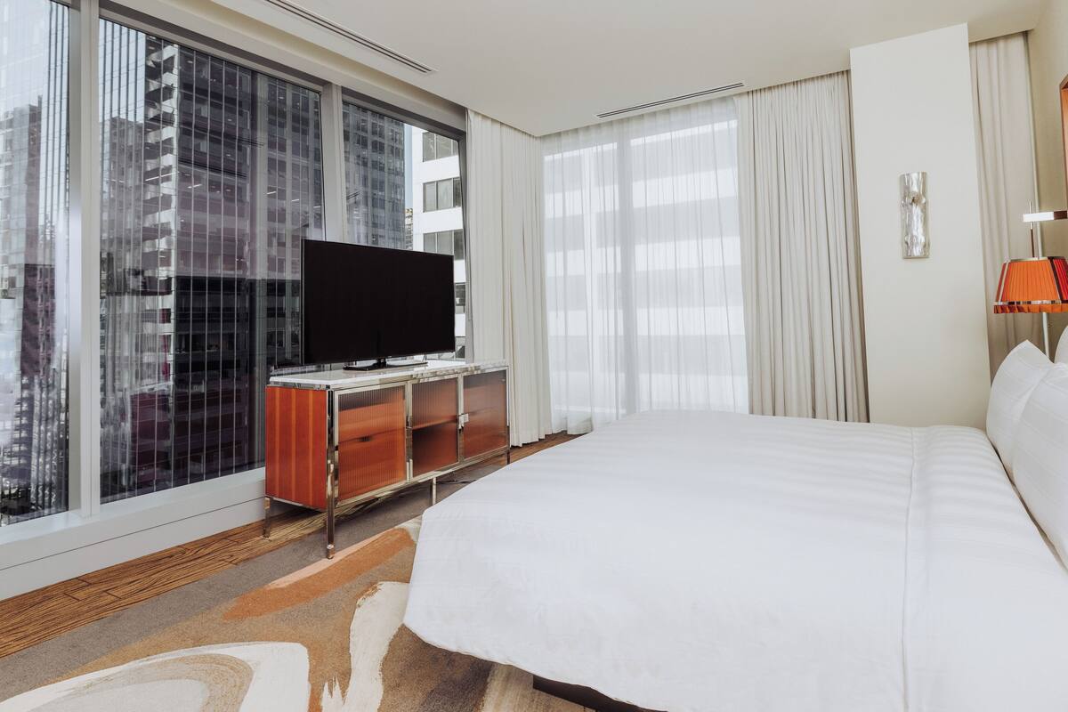 Quarto de hotel com uma cama grande, uma TV sobre um móvel de madeira e vista para a cidade através de janelas do chão ao teto.
