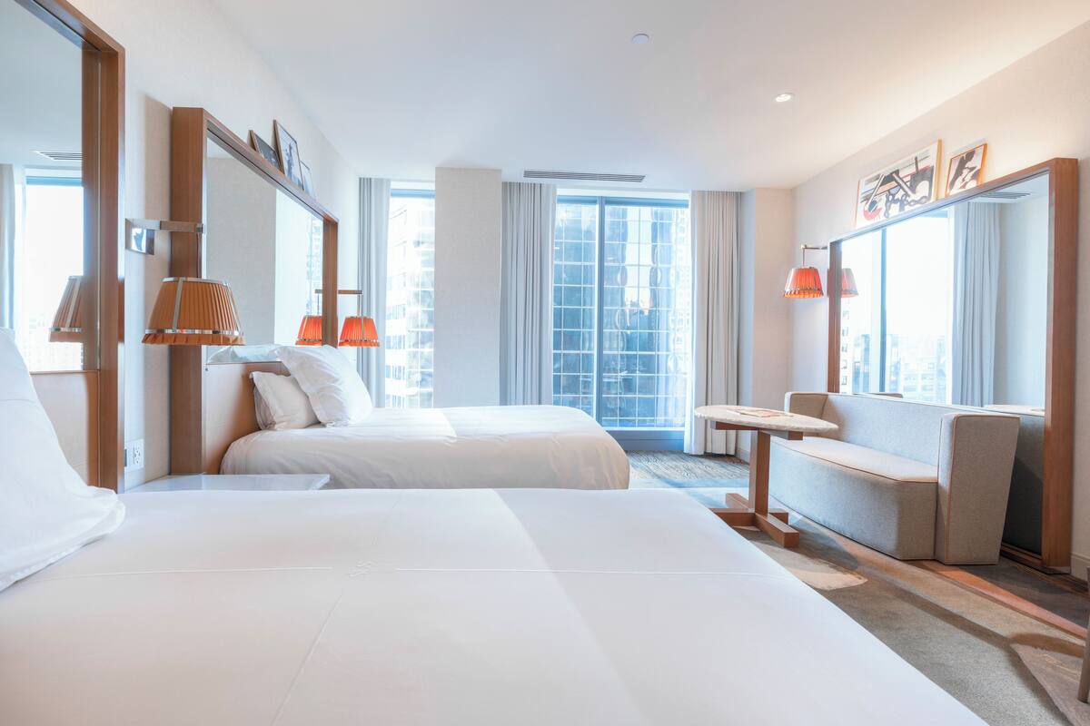 Quarto de hotel com duas camas, uma mesinha, um sofá e grandes janelas com vista para a cidade.