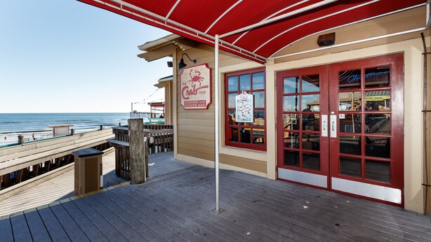 Um deck de madeira com toldo vermelho, uma placa com a inscrição 'The Crab Trap Seafood and Oyster Bar' e vista para o oceano.