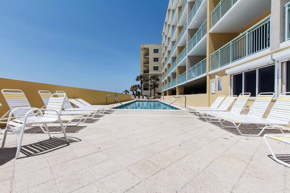 Área de piscina com espreguiçadeiras e um edifício ao fundo.
