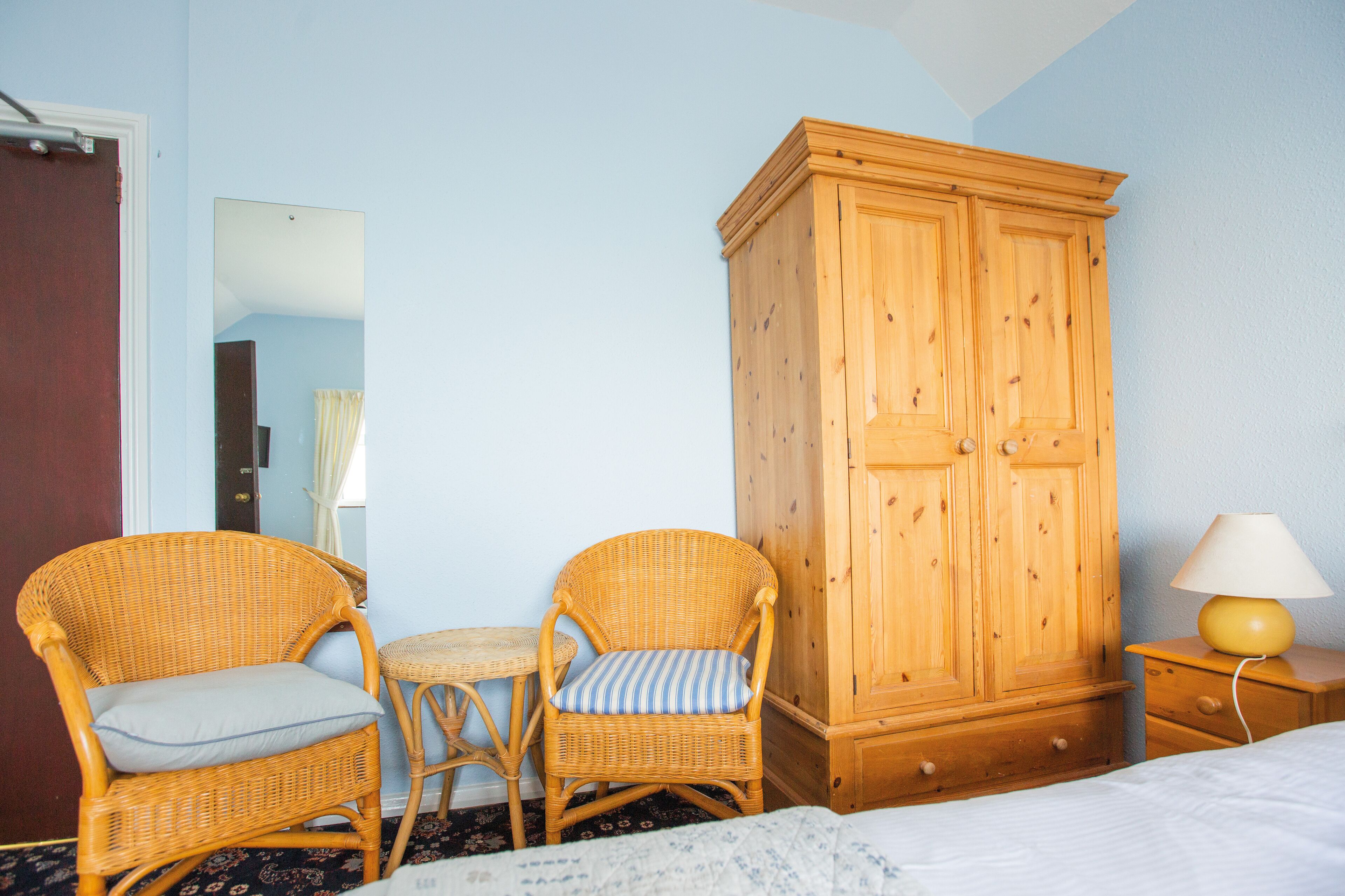 Quarto com cama, duas cadeiras de vime, guarda-roupa de madeira, mesa pequena e abajur.