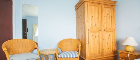Quarto com cama, duas cadeiras de vime, guarda-roupa de madeira, mesa pequena e abajur.