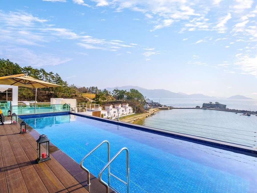 Piscina com deck de madeira, com vista para um corpo de água e montanhas ao longe.