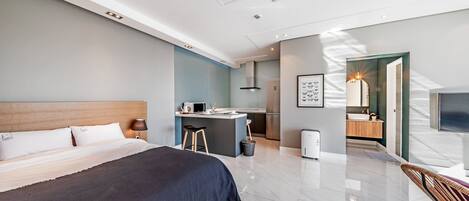 Quarto de hotel moderno com uma cama grande, uma kitchenette pequena, casa de banho com espelho e lavatório, e zona de estar com uma cadeira.