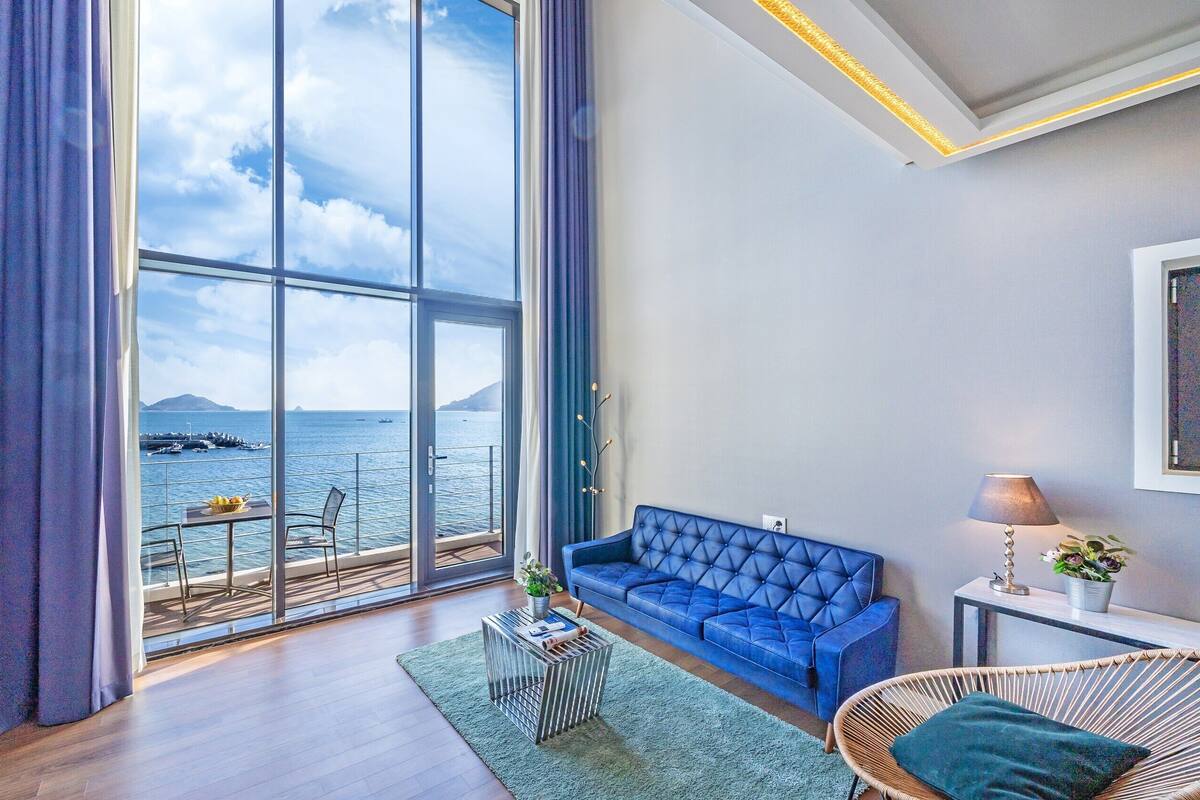 Uma sala de estar moderna com uma grande janela vista para o mar, um sofá azul, uma poltrona de vime e uma mesa pequena com abajur.
