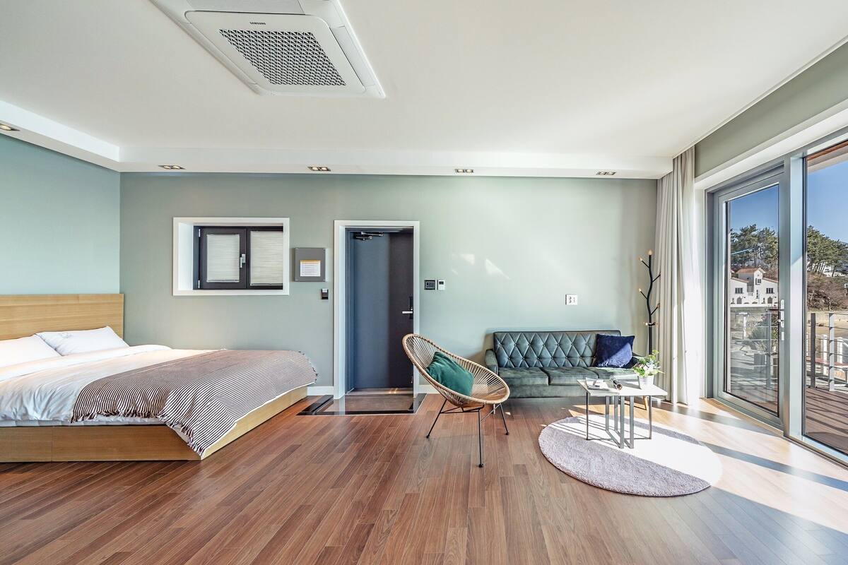 Quarto moderno com cama, área de estar com poltrona e sofá, mesa pequena e grande janela com vista para o mar.