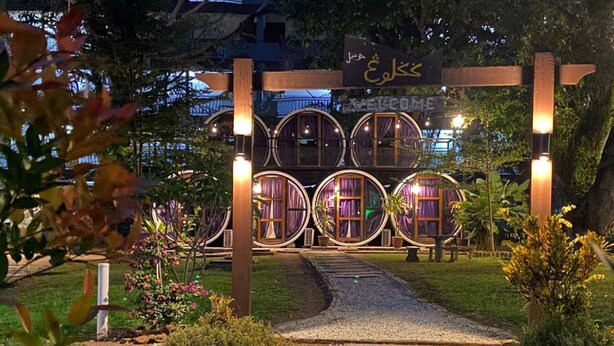 Entrada para um jardim, com um caminho que leva a estruturas circulares com cortinas, uma placa de boas-vindas e uma placa com texto em árabe.