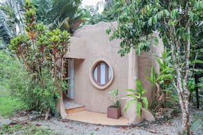 Um edifício singular, integrado à natureza, com uma janela circular, rodeado por vegetação exuberante e um caminho de cascalho.