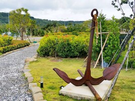 Um jardim com uma escultura de âncora enferrujada ao centro, cercado por sebes bem cuidadas e um caminho de cascalho.