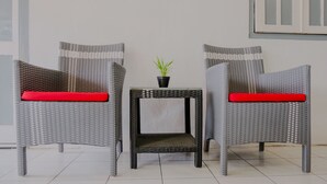 Duas cadeiras de vime com almofadas vermelhas, uma pequena mesa com uma planta e uma janela com vidros foscos.