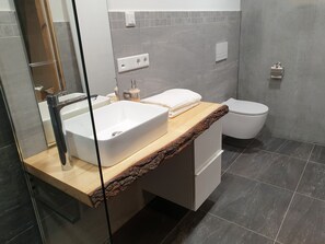 Uma casa de banho moderna com um lavatório de madeira, uma sanita, uma pia branca e um chuveiro com parede de vidro.