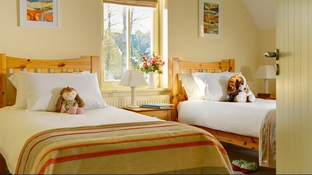 Quarto de hotel com duas camas de madeira, uma janela com vista para árvores e uma pequena mesa com uma luminária e livros.