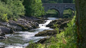 Um rio serpenteando sob uma ponte de pedra, cercado por vegetação exuberante.