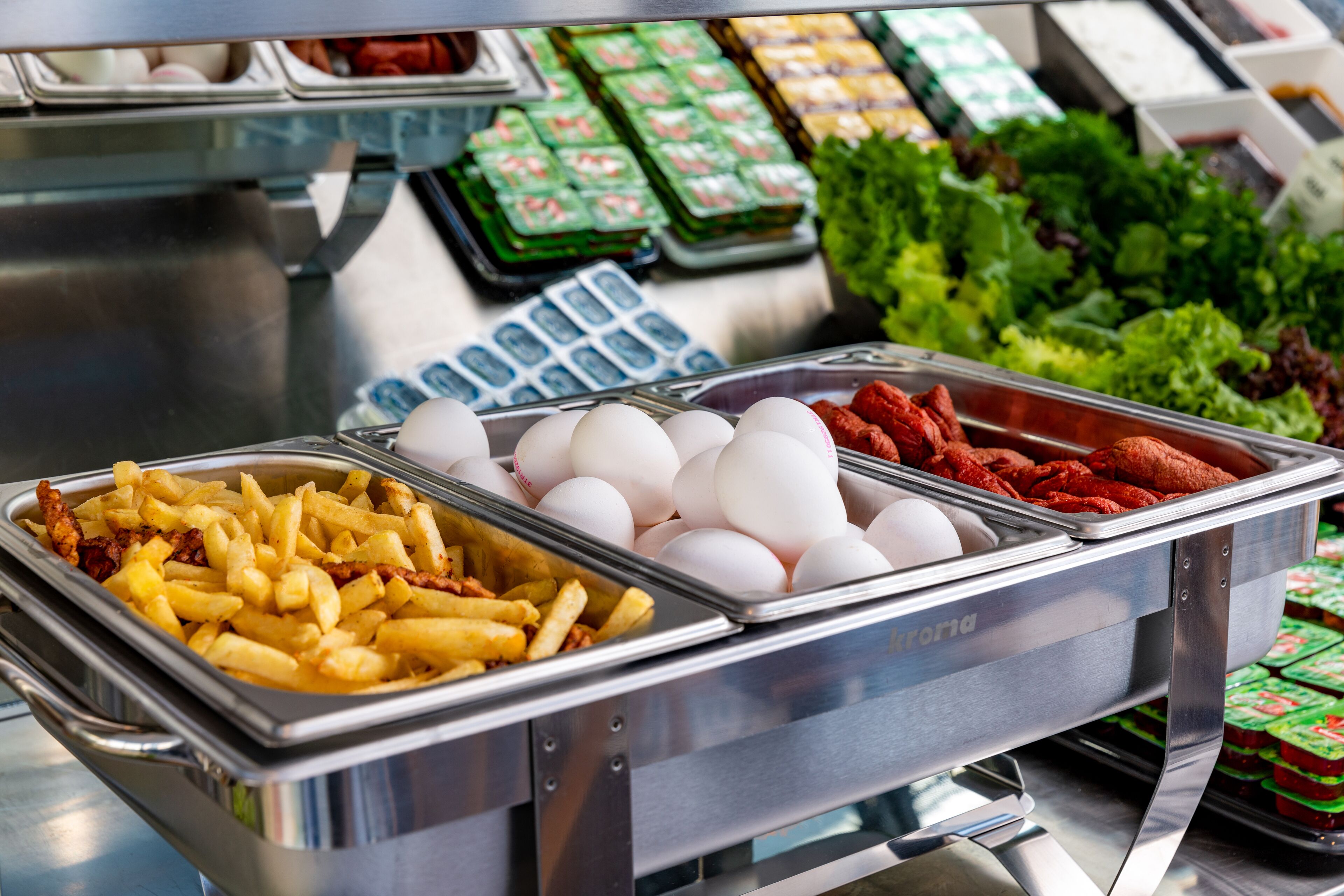 Buffet com bandejas de comida, incluindo ovos, batatas fritas e carne vermelha.