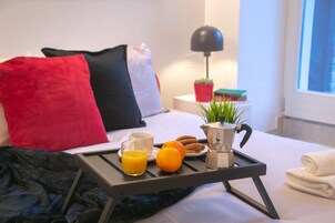 Cama arrumada com travesseiros vermelhos e pretos, mesa de cabeceira com abajur, uma pequena planta e uma bandeja com suco de laranja, biscoitos e uma cafeteira.