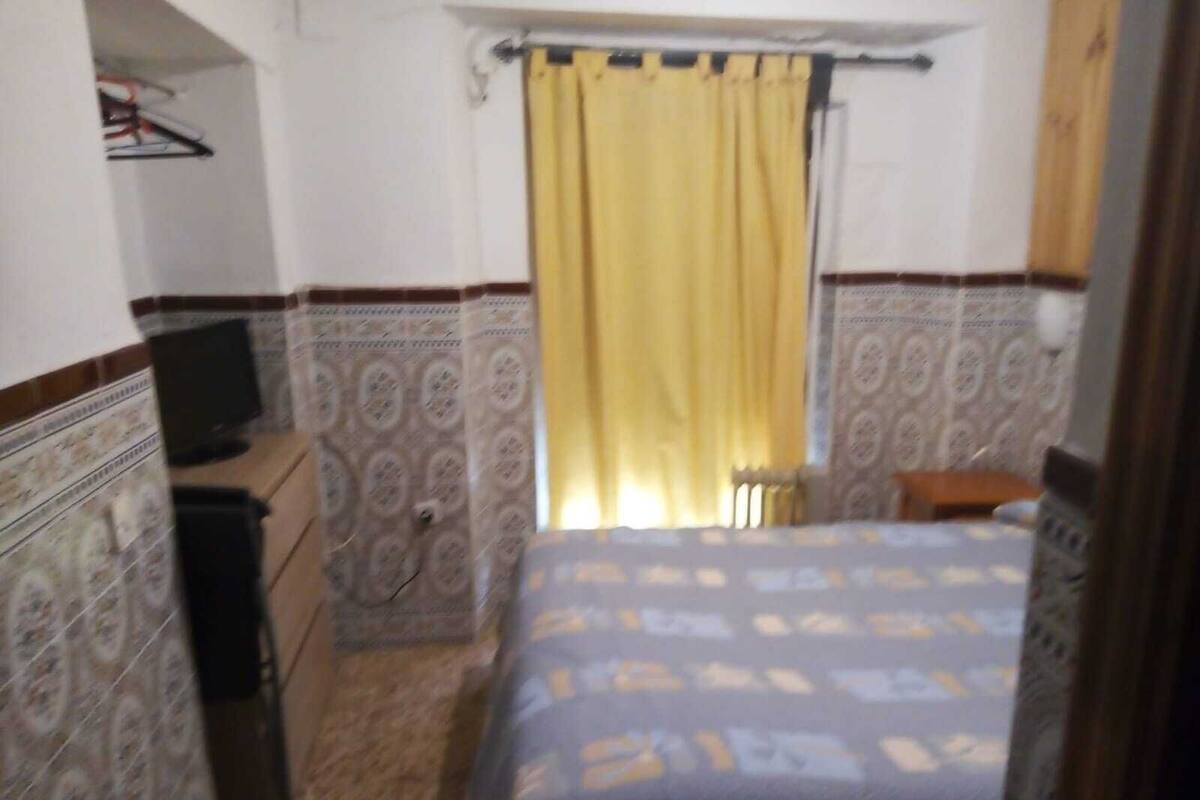 Um quarto pequeno e estreito, com uma cama, uma escrivaninha e uma televisão.
