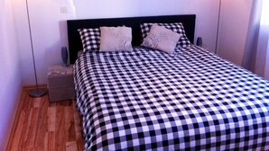 Uma cama bem arrumada com colcha xadrez, duas luminárias de cabeceira, uma mesinha de cabeceira pequena e piso de madeira.