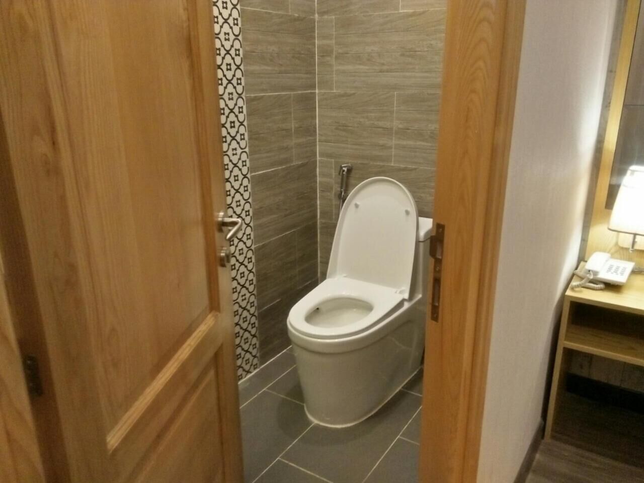 Uma porta de madeira dá acesso a um banheiro com vaso sanitário, cortina de chuveiro estampada e mesa de cabeceira com abajur e telefone.