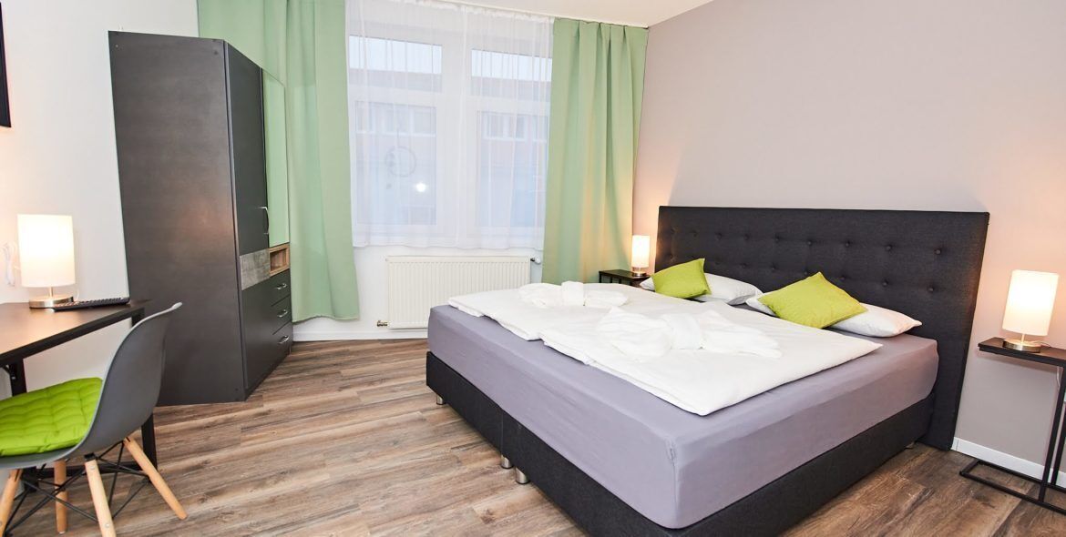 Quarto de hotel com uma cama, uma escrivaninha, uma cadeira e um guarda-roupa.